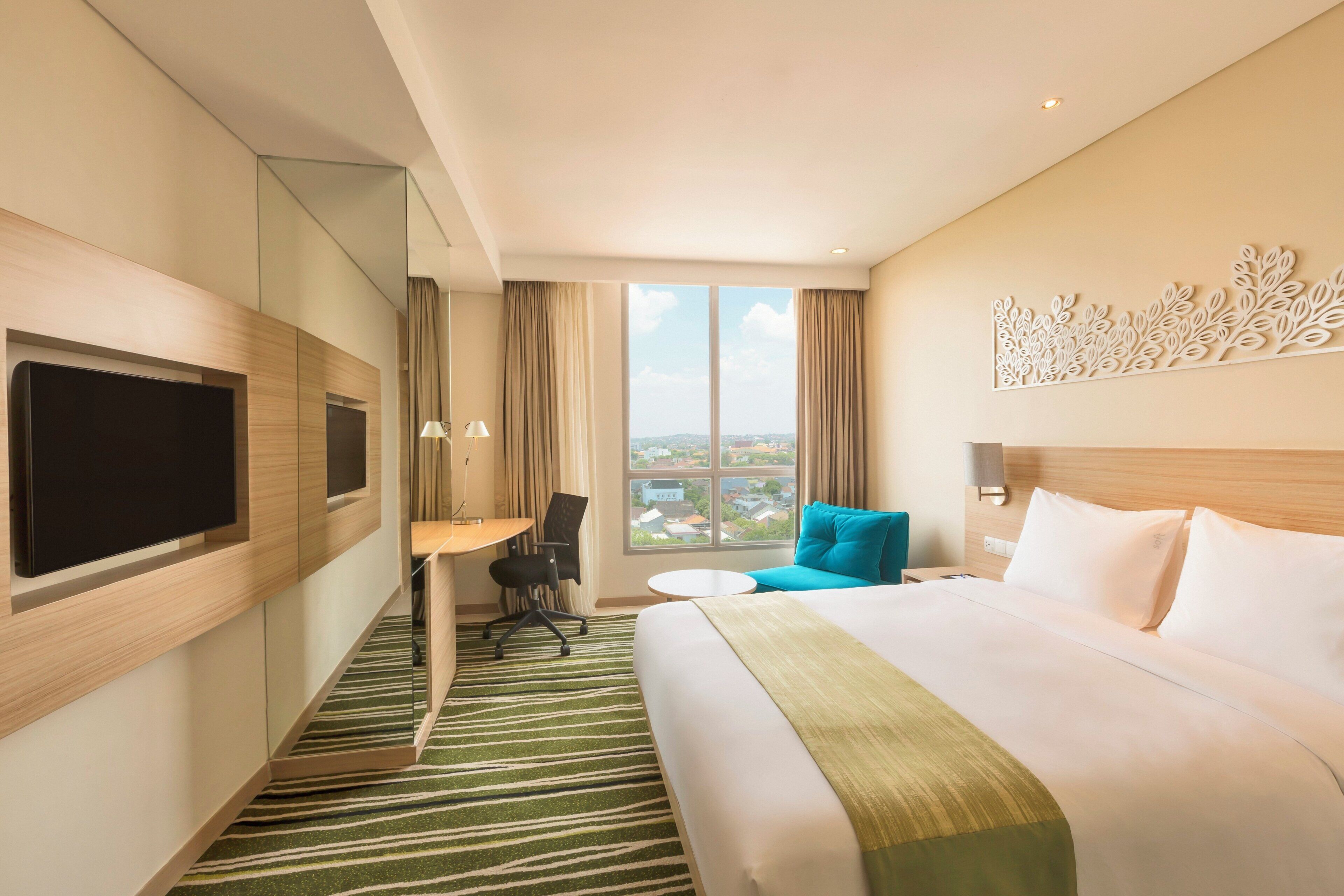 Quarto de hotel com cama, escrivaninha, cadeira, televisão, janela com vista e um detalhe decorativo na parede.