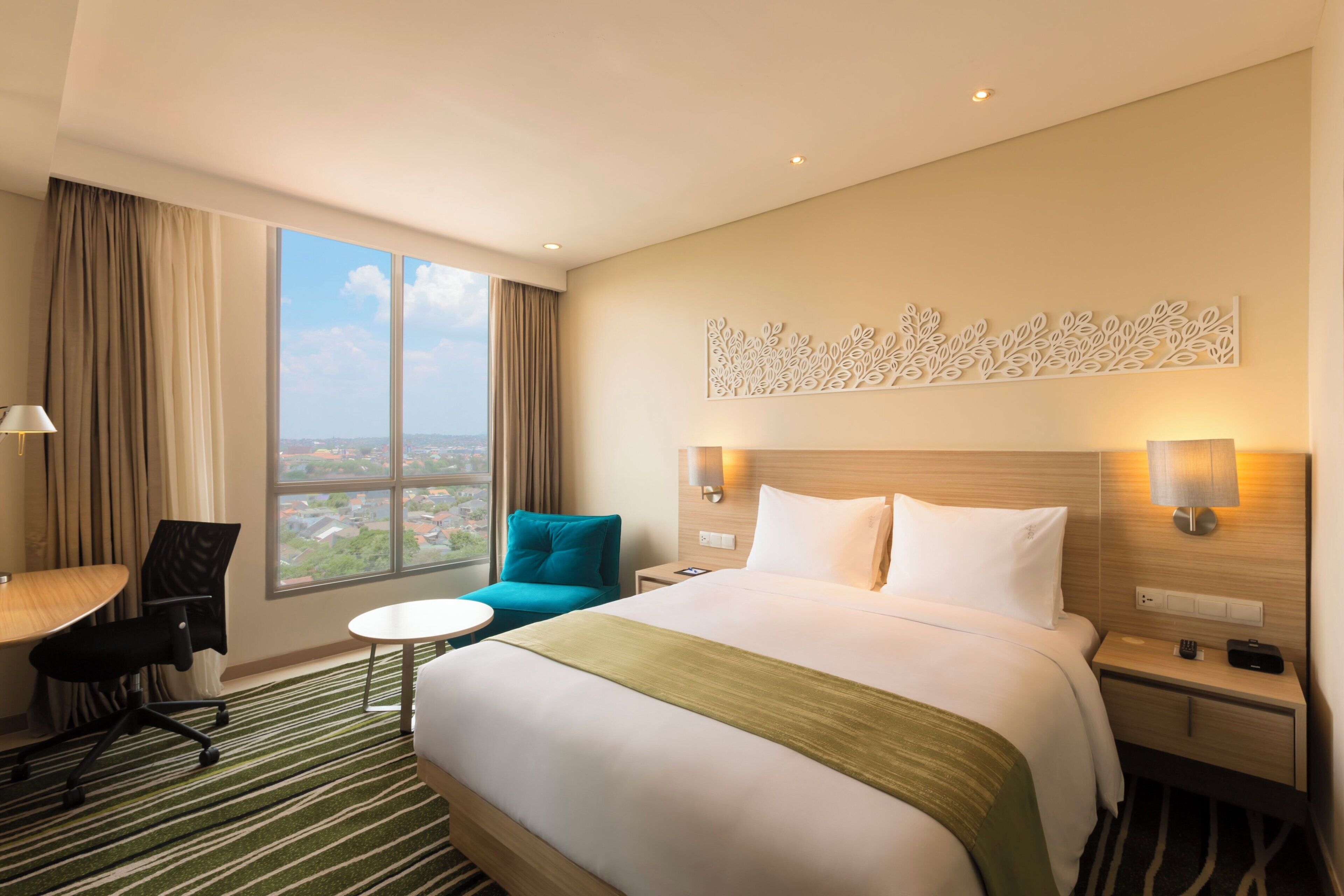 Quarto de hotel com uma cama grande, uma escrivaninha com cadeira, uma mesinha e vista para a cidade.