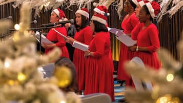 Um grupo de pessoas vestidas de vermelho e com gorros de Papai Noel cantando em um espaço interno decorado.