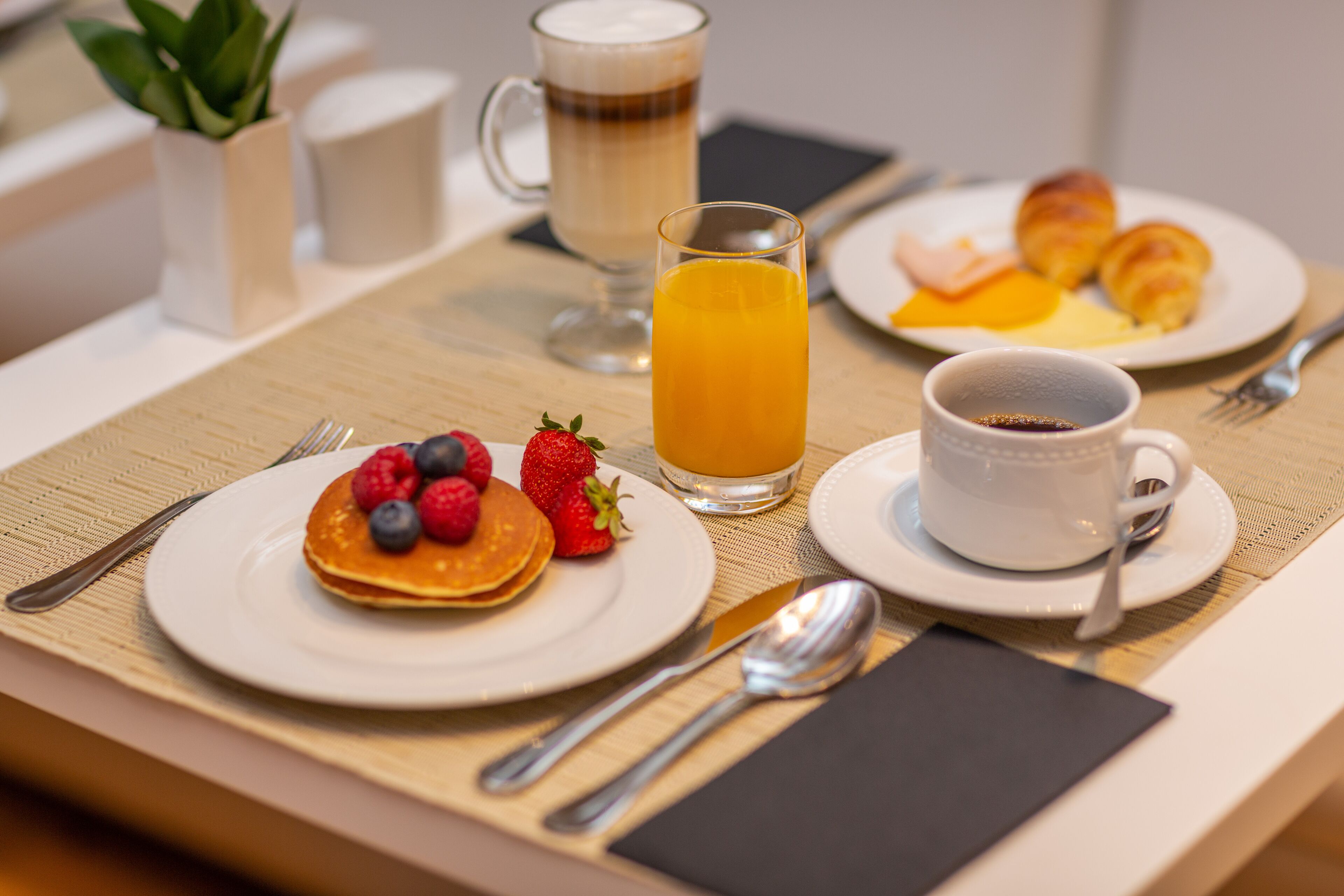 Mesa de café da manhã com panquecas cobertas de frutas vermelhas, um copo de suco de laranja, uma xícara de café, croissants e um prato de ovos.