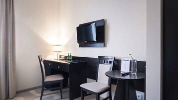 Quarto de hotel com escrivaninha, duas cadeiras, televisão de tela plana, abajur, pequena mesa com cardápio e um vaso com uma flor.