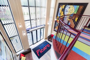 Um hall de hotel com um tapete colorido, um capacho preto com a inscrição 'HOTEL Picar', um sofá de couro marrom e uma grande pintura abstrata.