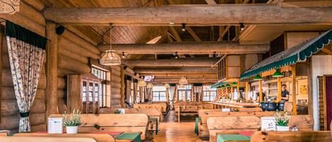 Um restaurante em estilo cabana de tronco, com bancos e mesas de madeira, área de bar e teto alto com vigas aparentes.