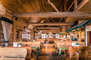 Um restaurante em estilo cabana de tronco, com bancos e mesas de madeira, área de bar e teto alto com vigas aparentes.