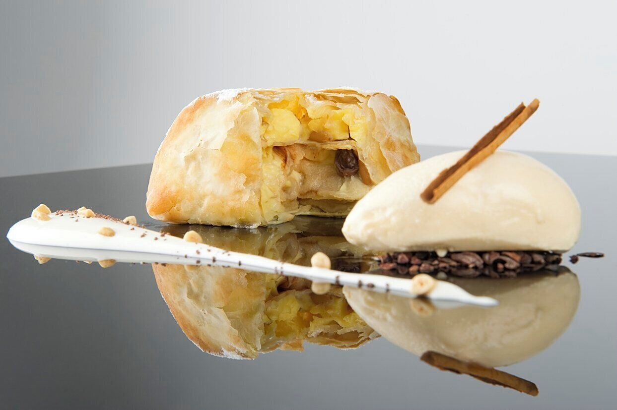 Um pastel com crosta dourada, recheio cremoso branco e camada de chocolate, acompanhado de uma colher e um palito pequeno.