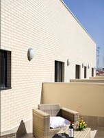 Um edifício moderno com fachada de tijolos brancos, uma varanda com sofá de vime, uma mesa com garrafa e copos, e um céu azul límpido.