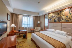 Quarto de hotel com uma cama grande, uma escrivaninha com luminária, uma cadeira, uma televisão e decoração na parede.