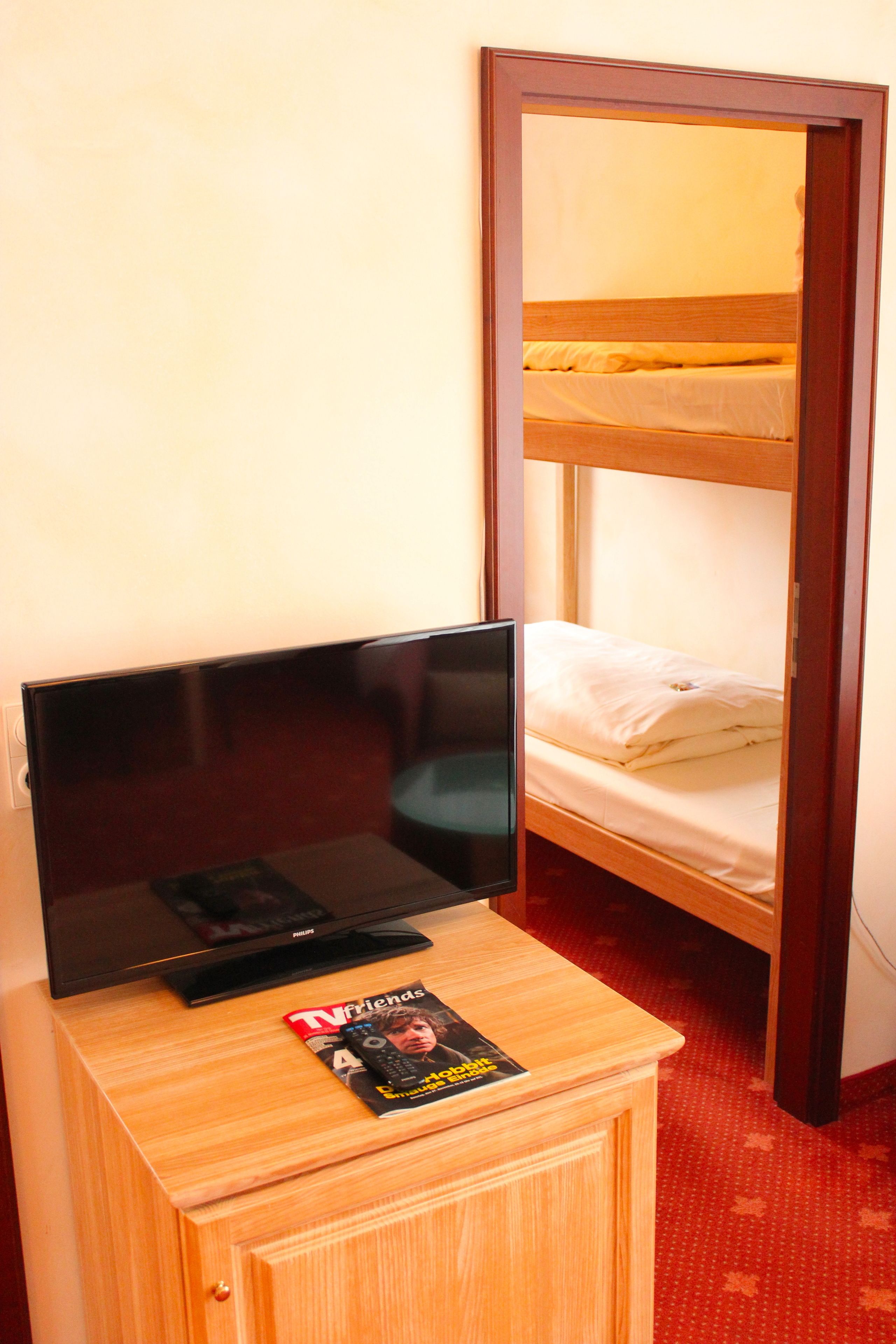 Quarto de hotel com mesa de cabeceira de madeira, televisão e uma revista.