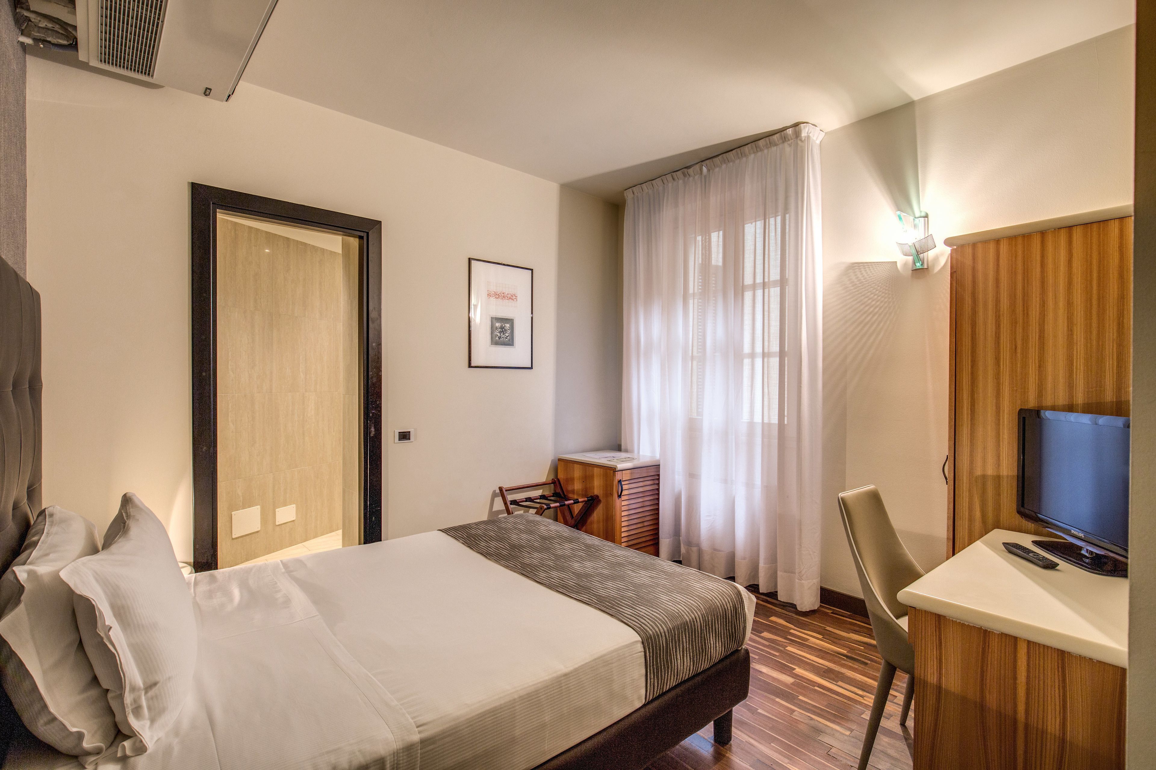 Quarto de hotel com cama, escrivaninha com televisão, cadeira, guarda-roupa e um quadro na parede.