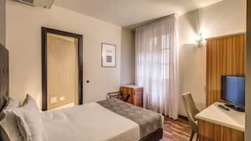 Quarto de hotel com cama, escrivaninha com televisão, cadeira, guarda-roupa e um quadro na parede.