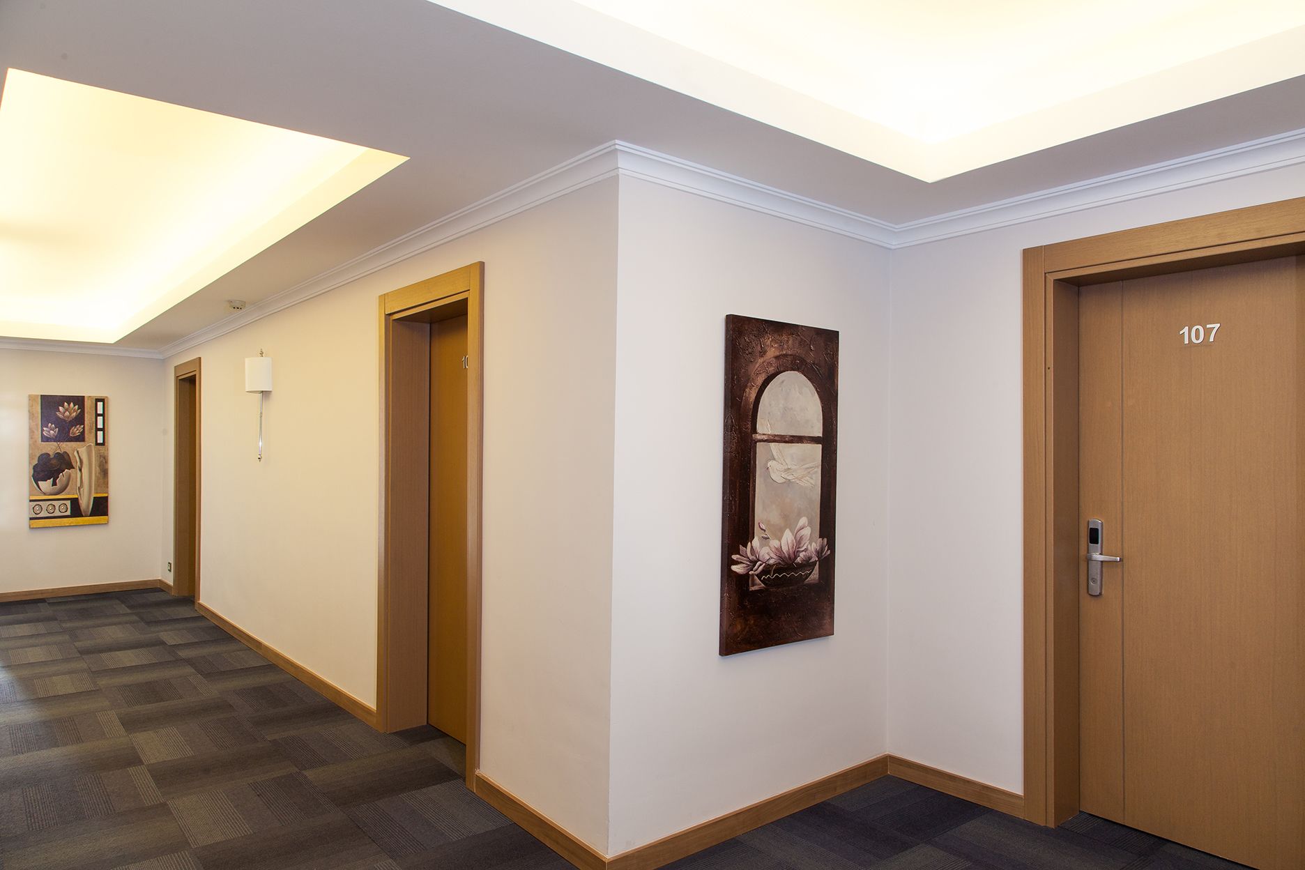 Um corredor de hotel com uma porta de madeira com a placa 107, uma obra de arte na parede e um candeeiro no teto.