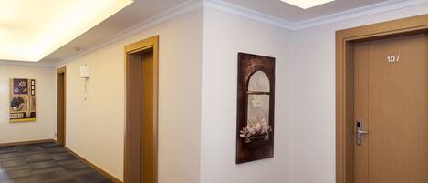 Um corredor de hotel com uma porta de madeira com a placa 107, uma obra de arte na parede e um candeeiro no teto.
