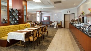 Um restaurante com área de bancos amarelos, várias mesas de jantar com cadeiras, um balcão de buffet e uma televisão na parede.