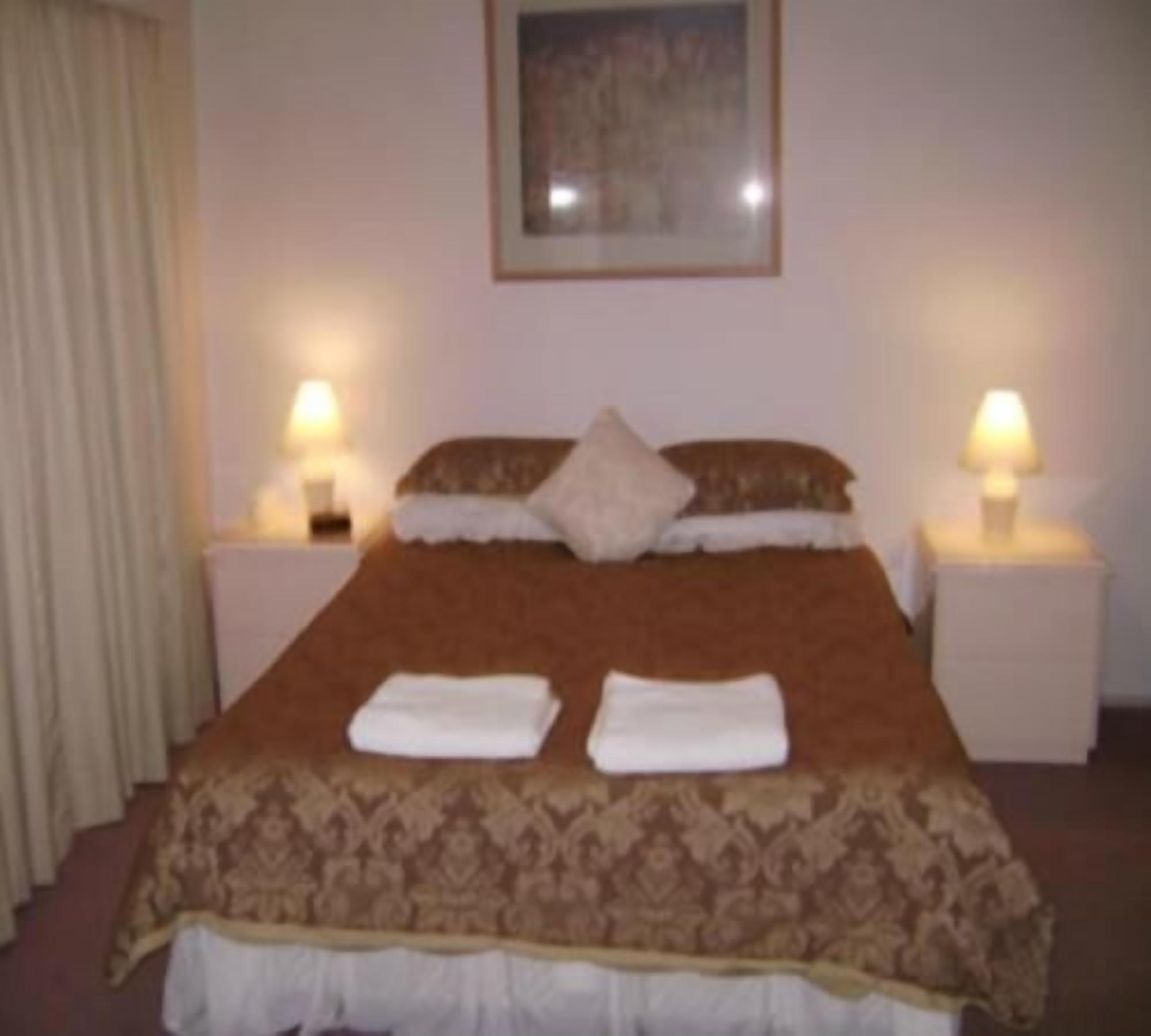 Cama bem arrumada com colcha estampada, dois travesseiros e duas toalhas dobradas. Duas luminárias de cabeceira em cada lado da cama.