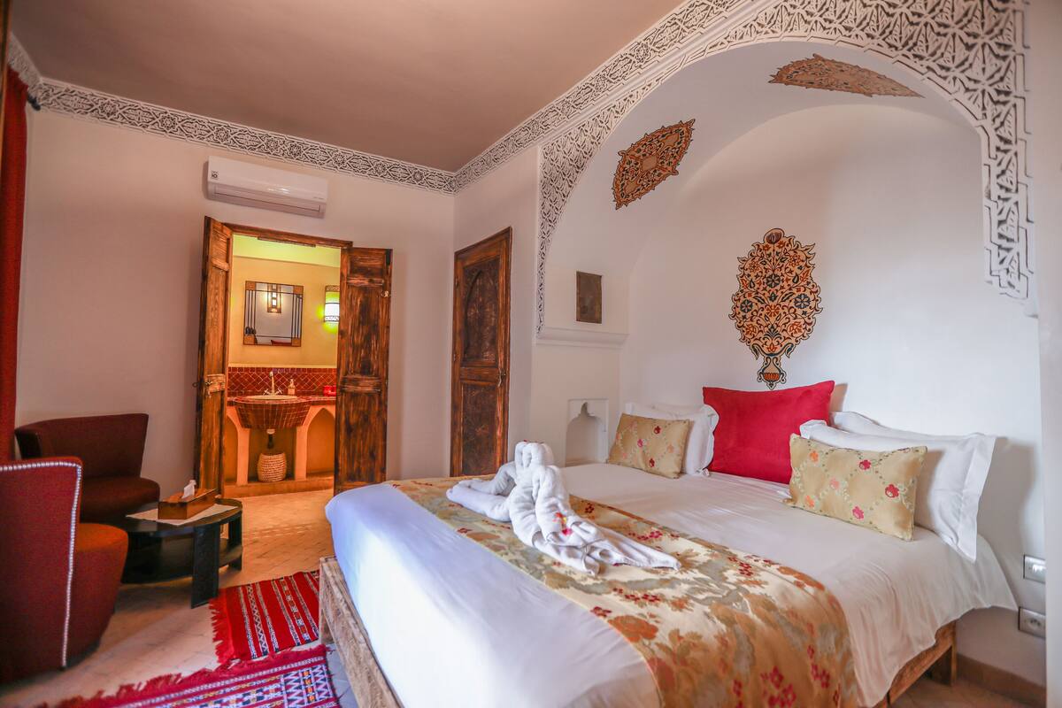 Quarto de hotel com cama, uma poltrona vermelha, uma mesinha e um banheiro visível através de uma porta aberta.