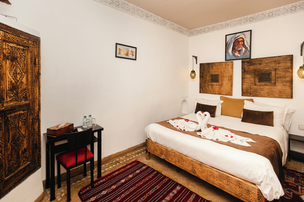 Quarto de hotel com uma cama grande, porta de madeira, uma mesinha com cadeira e um tapete vermelho.