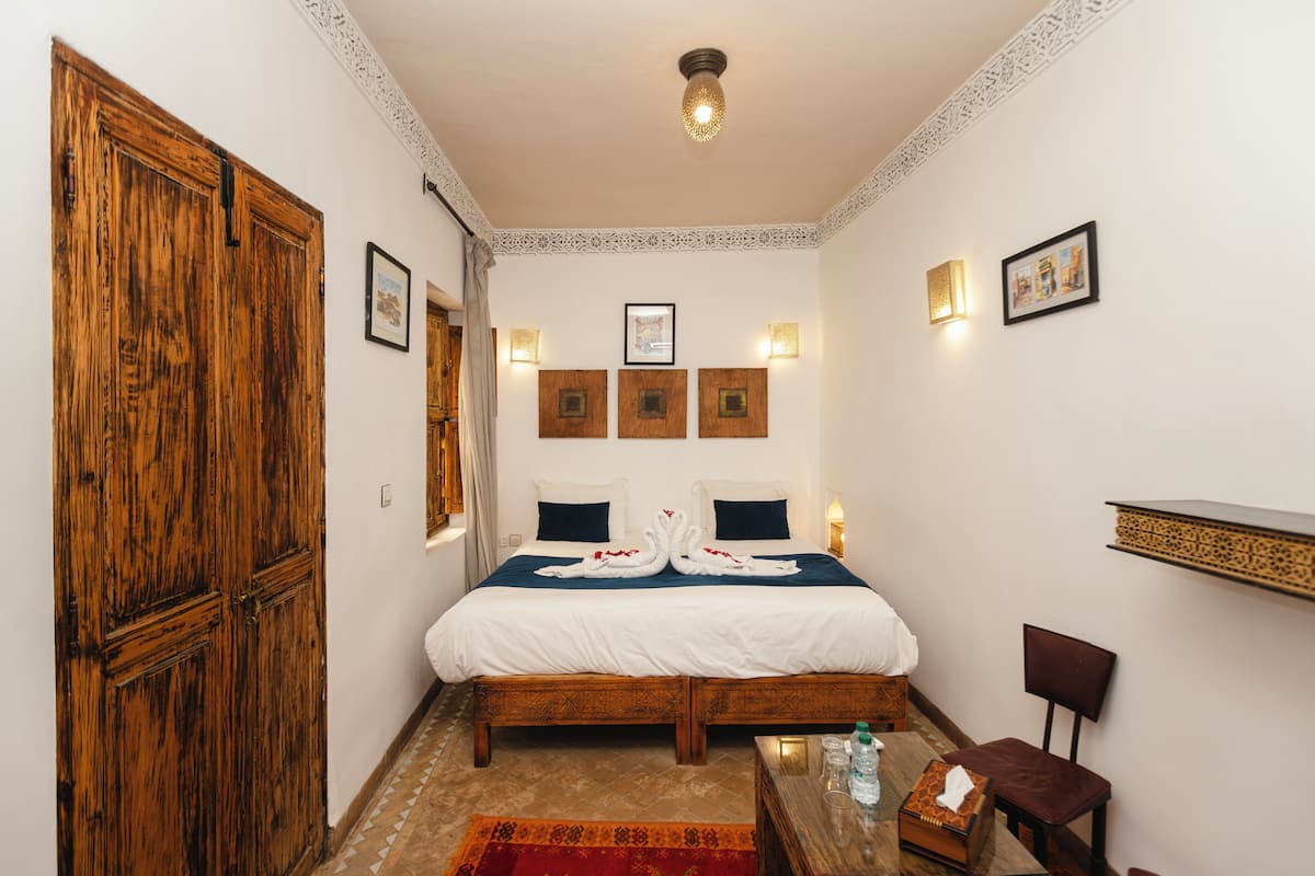 Quarto de hotel com uma cama de madeira, uma pequena mesa com uma garrafa e copos, uma cadeira e uma prateleira decorativa na parede.