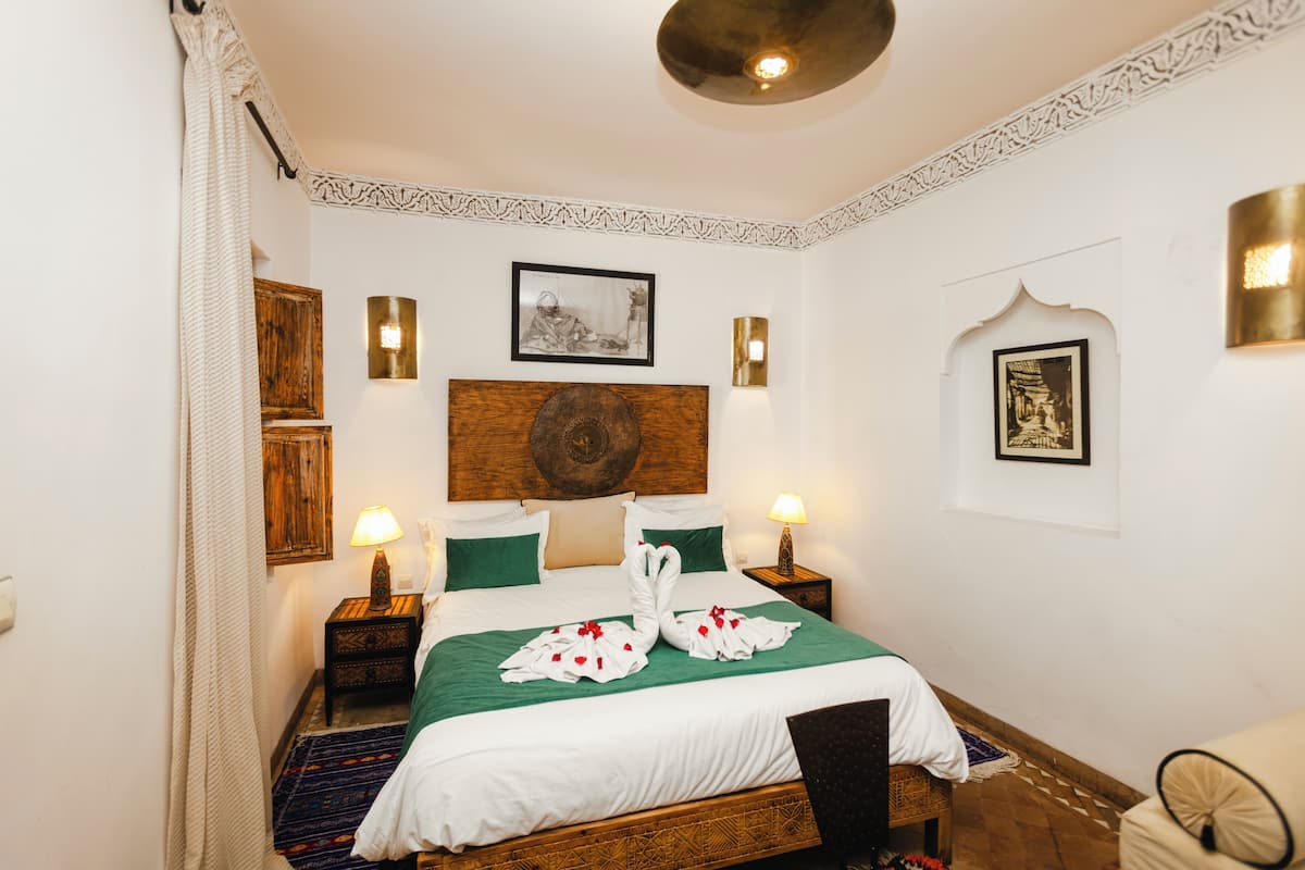 Uma cama bem arrumada com um edredom verde e lençóis brancos, duas luminárias fixadas na parede e uma moldura com uma imagem sobre a cama.