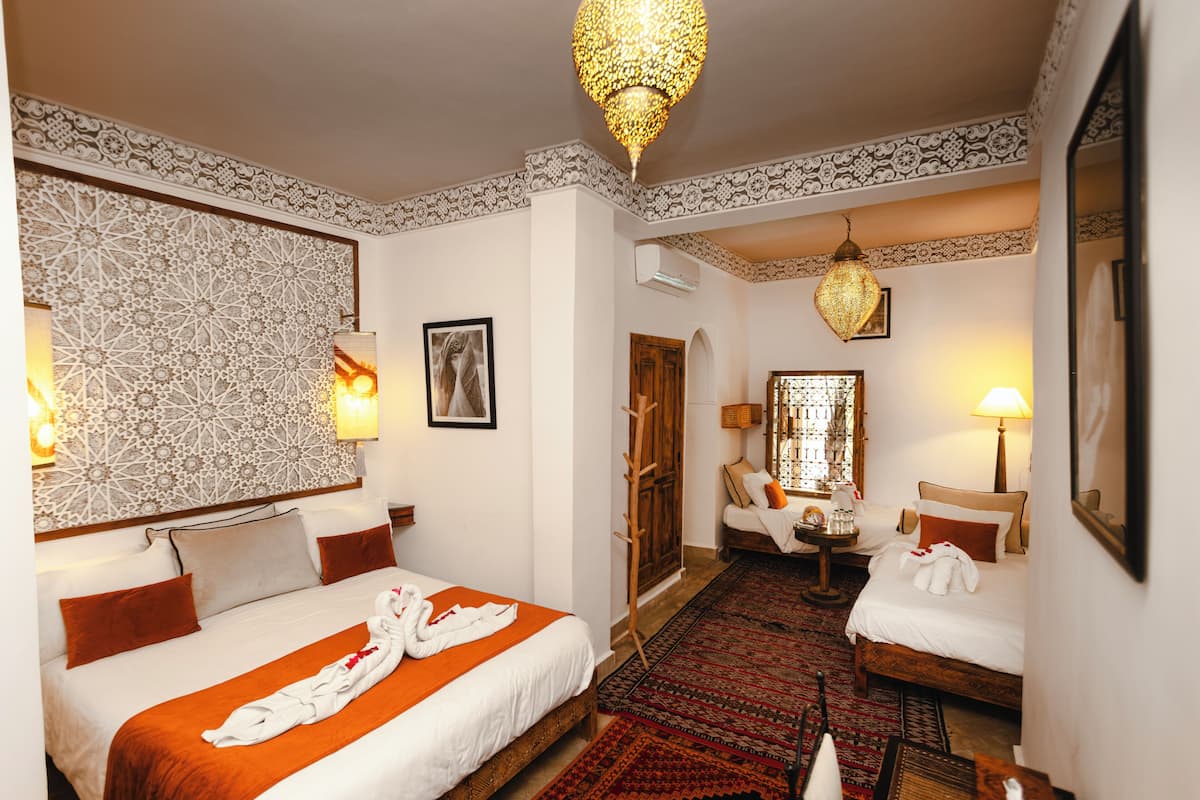 Quarto de hotel com uma cama grande, uma cama menor, área de estar com uma cadeira e mesa, e parede decorativa com padrões elaborados.