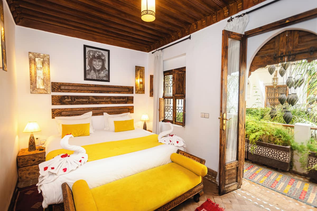 Quarto de hotel com cama, cabeceira de madeira, luminárias laterais e vista para um jardim através de uma porta aberta.