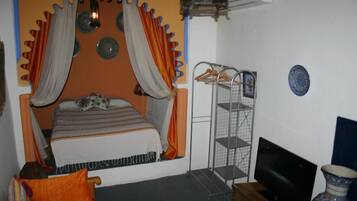Quarto com cama, cortinas, prateleira de metal, televisão e um vaso.