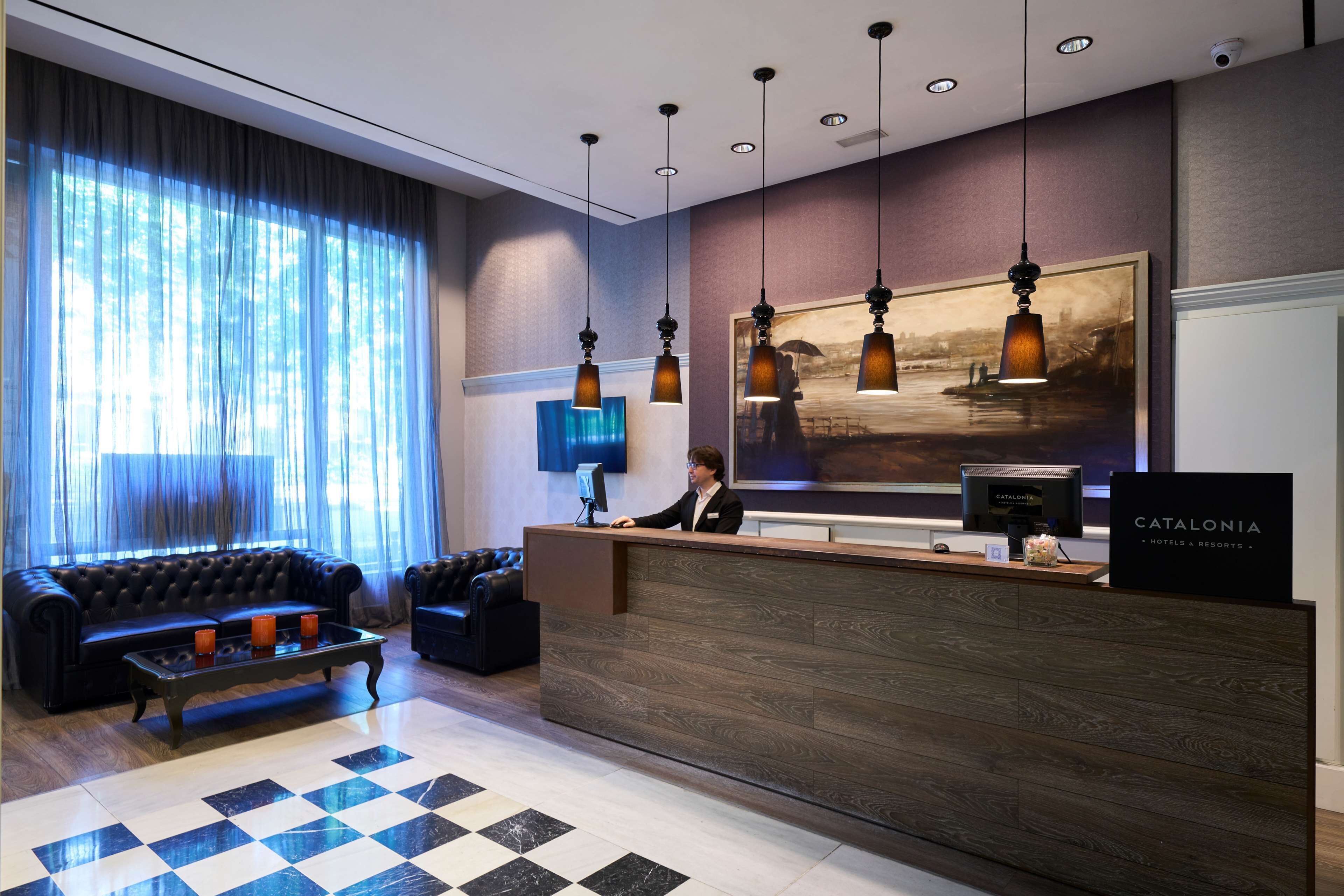 Uma área de recepção de hotel com balcão de madeira, sofá de couro preto, piso xadrez e luminárias pendentes.