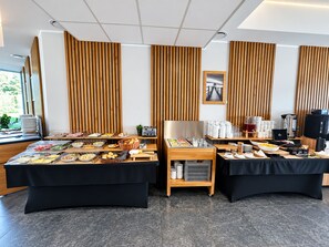 Disposição de buffet com diversos alimentos e um canto de café, com o fundo de ripas de madeira e um quadro emoldurado.