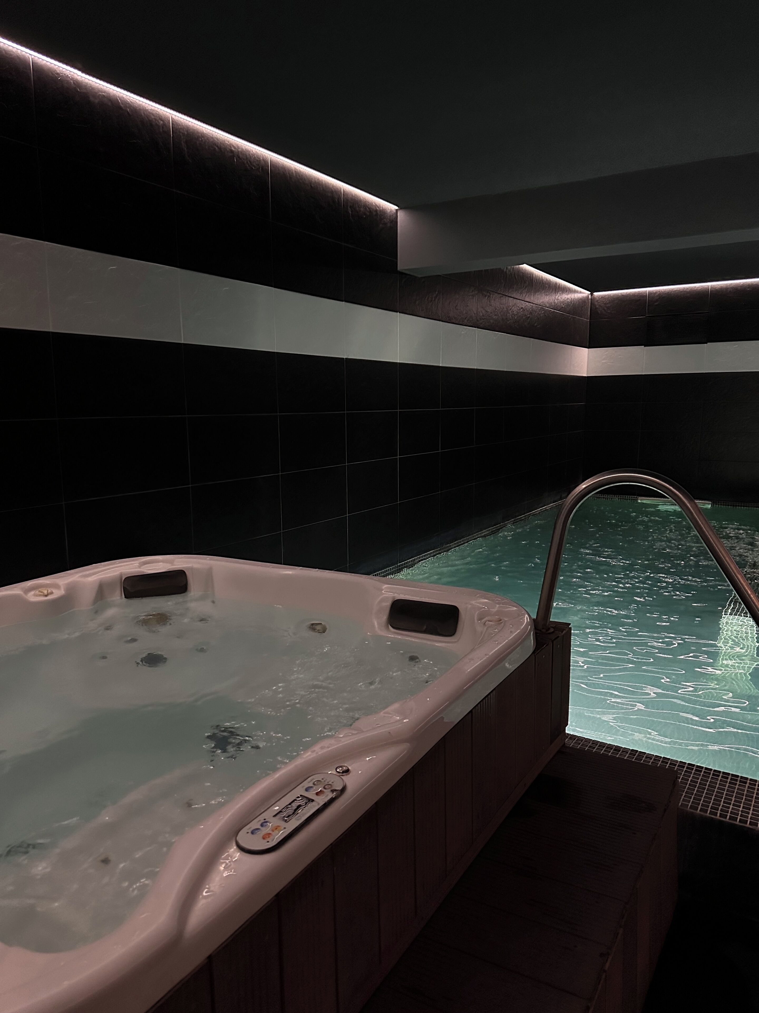 Uma banheira de hidromassagem com base de madeira, cercada por paredes revestidas de azulejos escuros e uma piscina transparente.