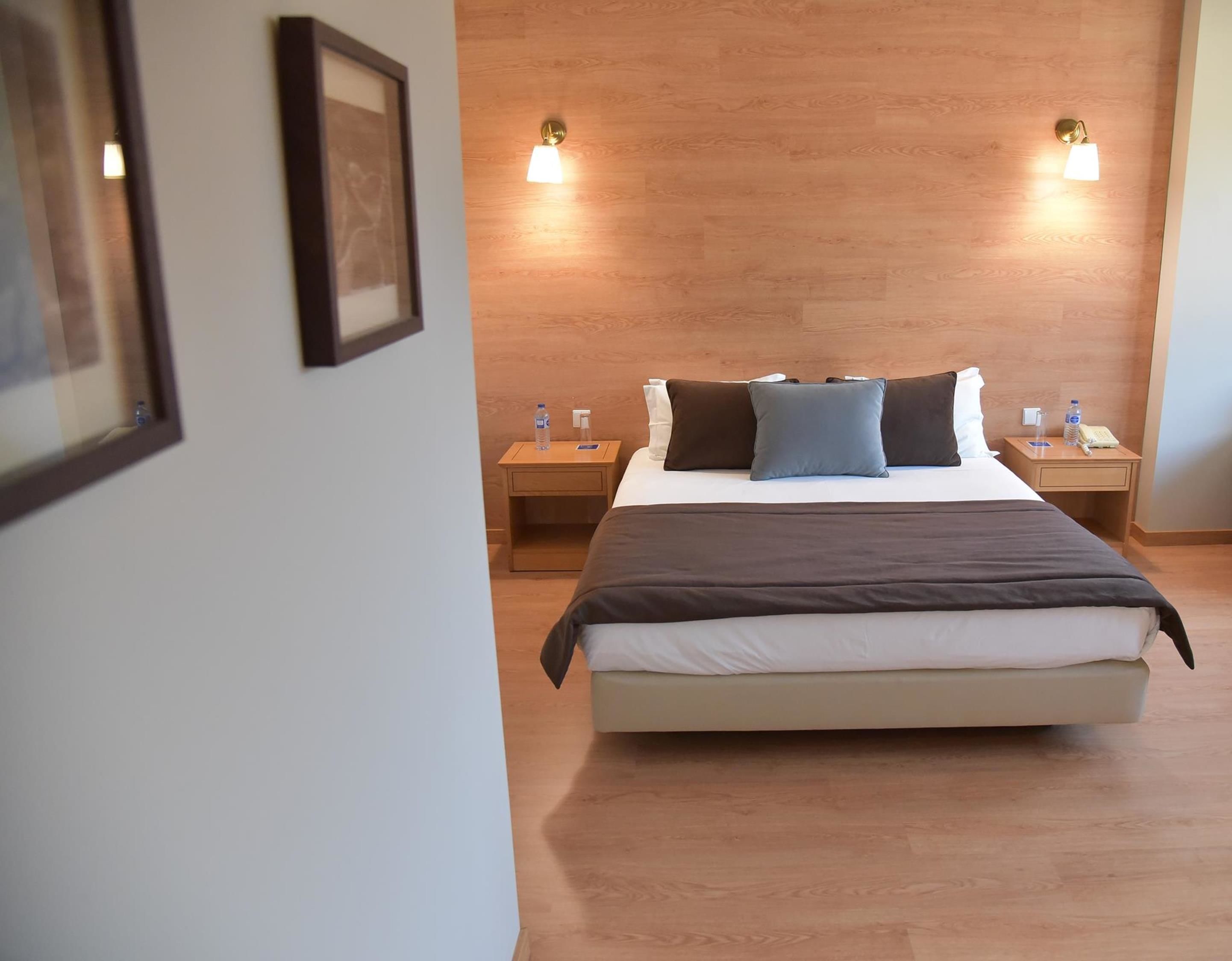 Quarto de hotel com uma cama grande, duas mesas de cabeceira, luminária fixada na parede e um quadro emoldurado.