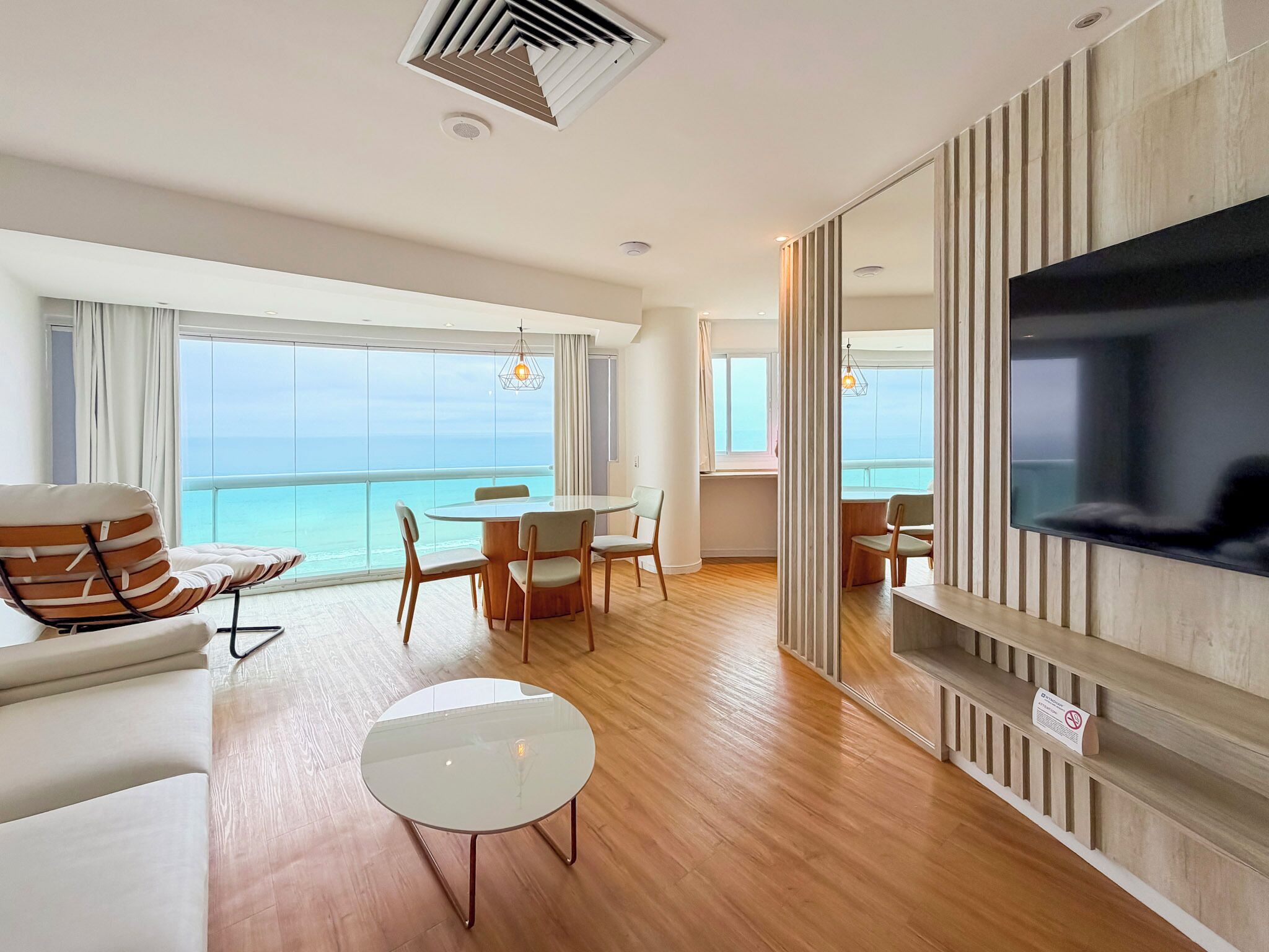 Uma sala de estar moderna com uma televisão grande, uma área de refeições e vista para o oceano.
