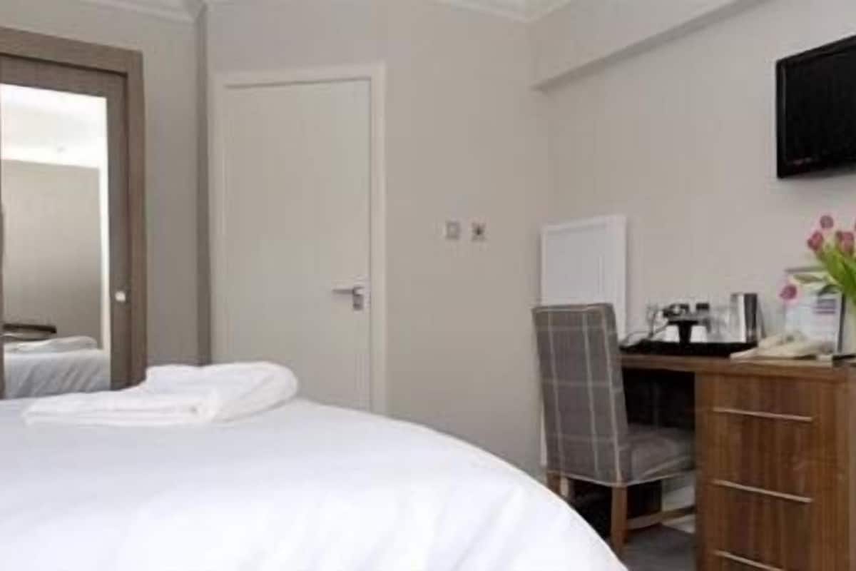 Quarto de hotel com uma cama, escrivaninha, cadeira, guarda-roupa e televisão na parede.