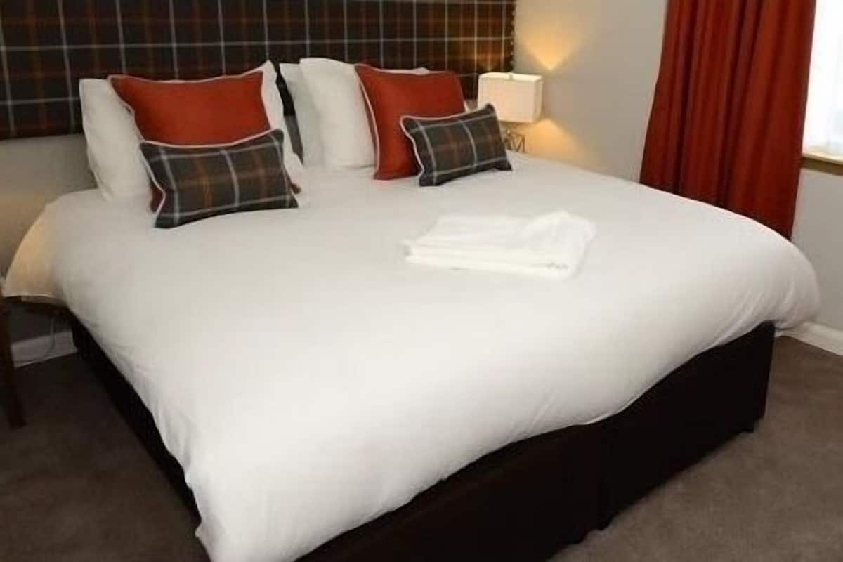 Uma cama bem arrumada com roupa de cama branca e almofadas xadrez, uma luminária de cabeceira e uma cortina vermelha.
