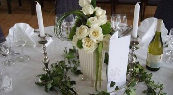 Mesa posta para um evento formal, com toalhas brancas, um vaso de flores brancas, velas e uma garrafa de vinho.
