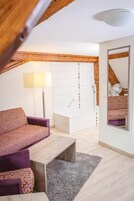 Um quarto pequeno e acolhedor com um sofá roxo, uma mesa de centro de madeira, uma banheira branca e um espelho refletindo um sofá estampado.