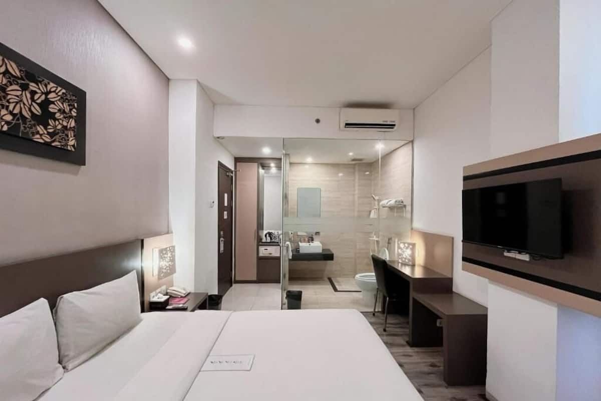 Quarto de hotel moderno com uma cama grande, uma escrivaninha, uma televisão e um banheiro visível por uma porta aberta.