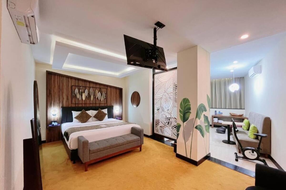 Quarto de hotel moderno com uma cama grande, escrivaninha, cadeira, esteira e televisão fixada na parede.