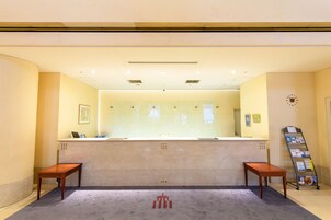 Uma área de recepção com balcão, duas bancadas de madeira, um tapete com um símbolo vermelho e um painel informativo fixado na parede.