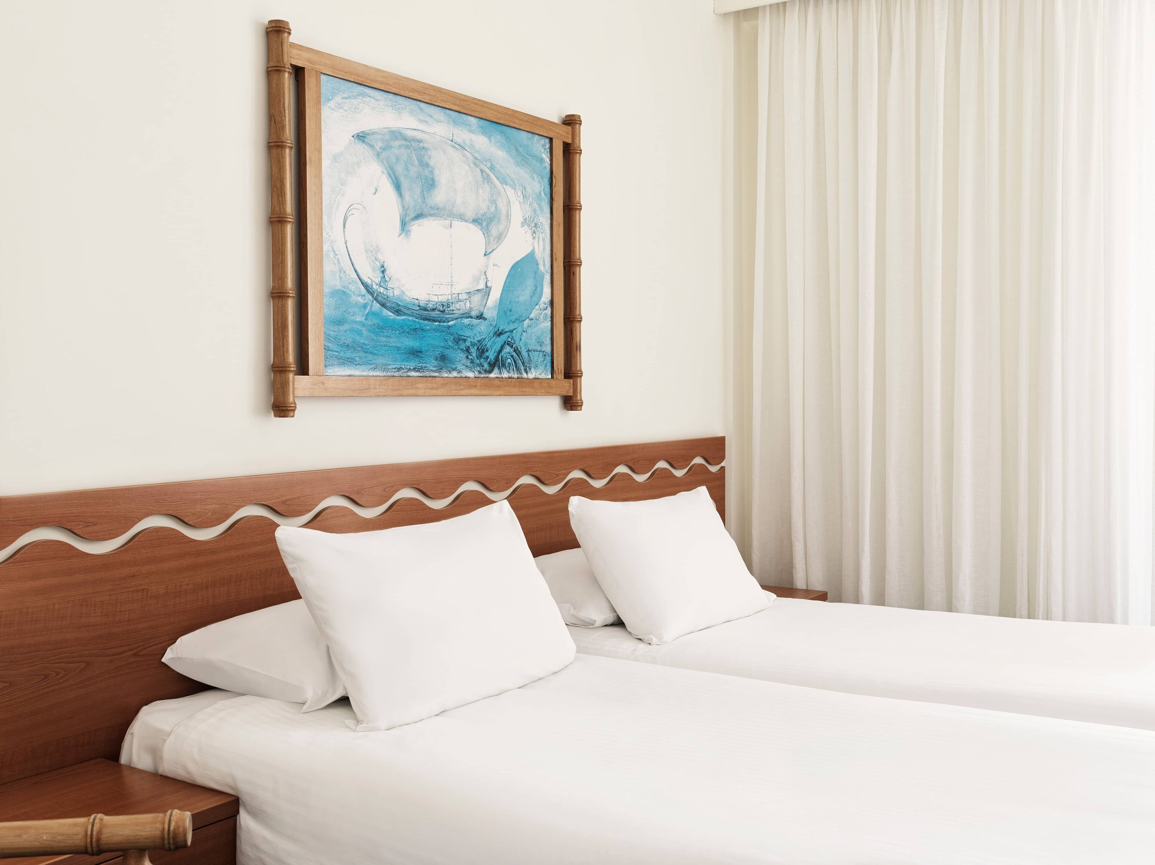 Quarto de hotel com cabeceira de madeira, duas almofadas brancas, um quadro abstrato grande e uma janela com cortinas brancas.
