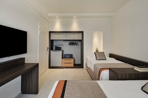 Quarto de hotel moderno com televisão de tela plana, uma cama, mesa de cabeceira, escrivaninha, armário e pequeno espaço de cozinha.