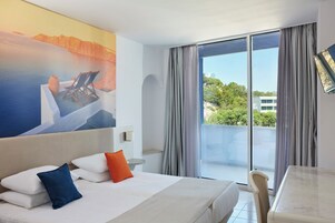 Quarto de hotel com cama, escrivaninha, cadeira, pintura de uma paisagem costeira e varanda com vista para áreas verdes e edifícios.