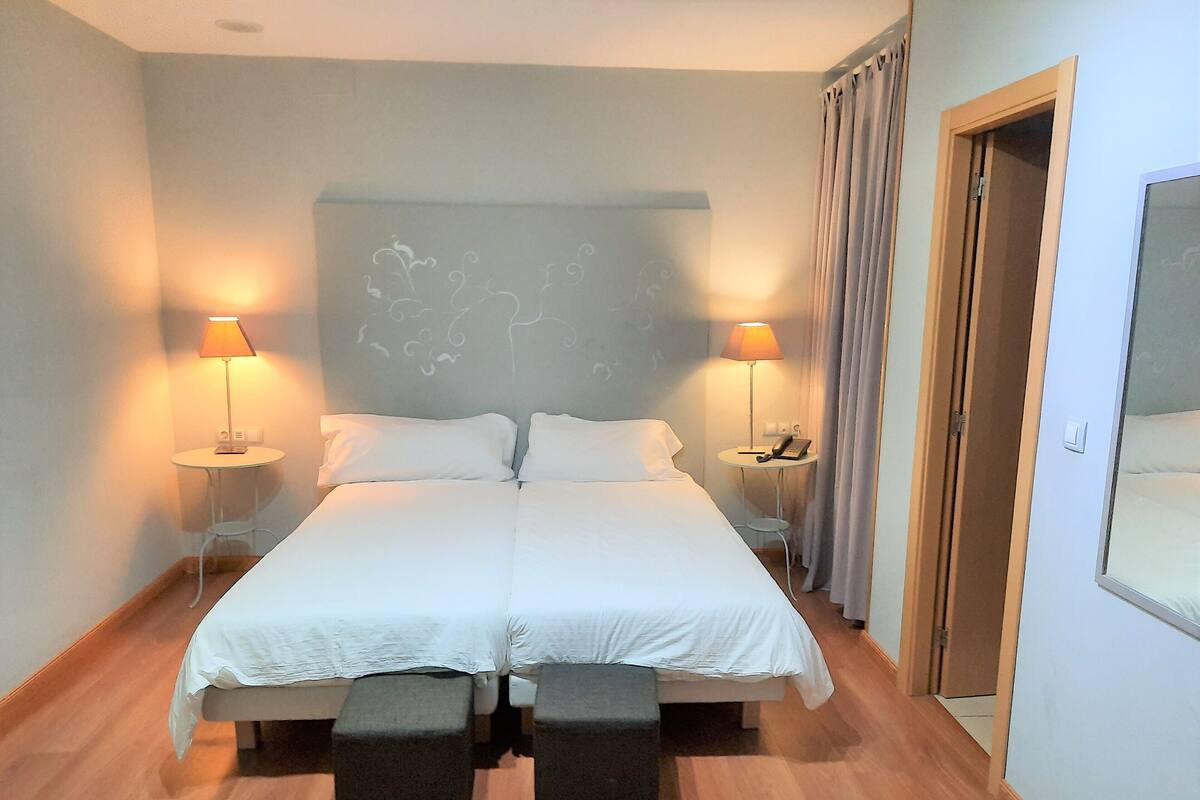 Uma cama de casal com roupa de cama branca, duas mesas de cabeceira com luminárias e uma cabeceira com padrão floral, tudo sobre um piso de madeira.