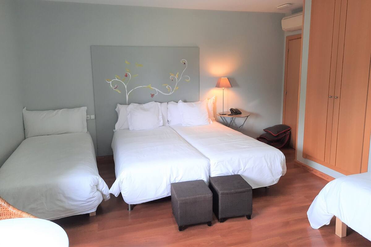 Quarto de hotel com duas camas, uma mesa de cabeceira com abajur, um guarda-roupa e um pequeno banquinho.