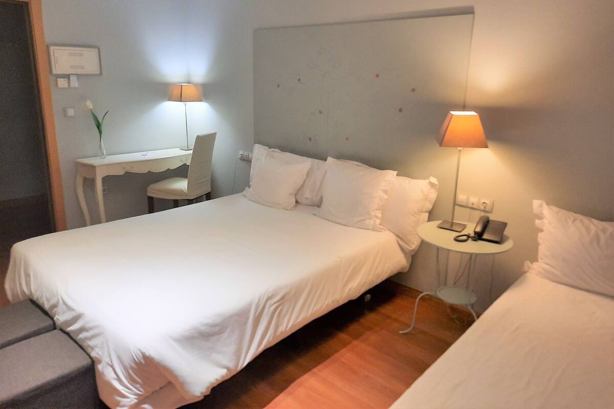 Quarto de hotel com cama, mesa de cabeceira com abajur, escrivaninha com cadeira e uma mesinha com telefone.