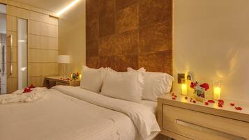 Quarto de hotel com cama grande, cabeceira de madeira, mesas de cabeceira e mesa de apoio com velas e porta-retrato.