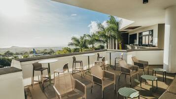 Terraço no telhado com cadeiras e mesas de vime, com vista para um horizonte urbano com palmeiras e colinas ao longe.