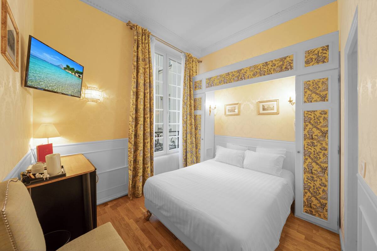 Quarto de hotel com cama, televisão na parede, mesa de cabeceira com abajur e janela com cortinas.