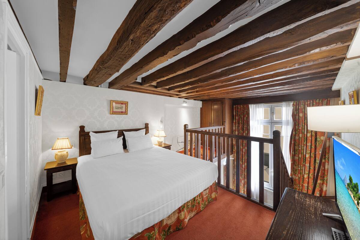 Quarto com cama grande, vigas de madeira no teto, mesa de cabeceira com abajur e televisão sobre uma mesa de madeira.
