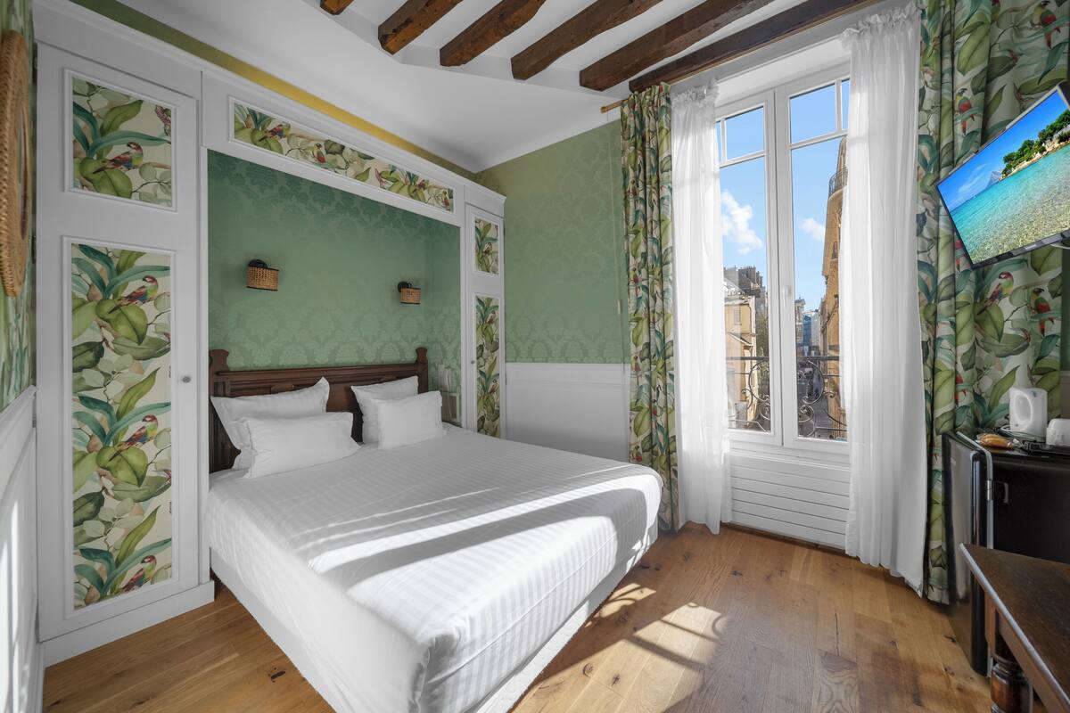 Quarto com cama, piso de madeira, papel de parede verde e janela com vista para edifícios.