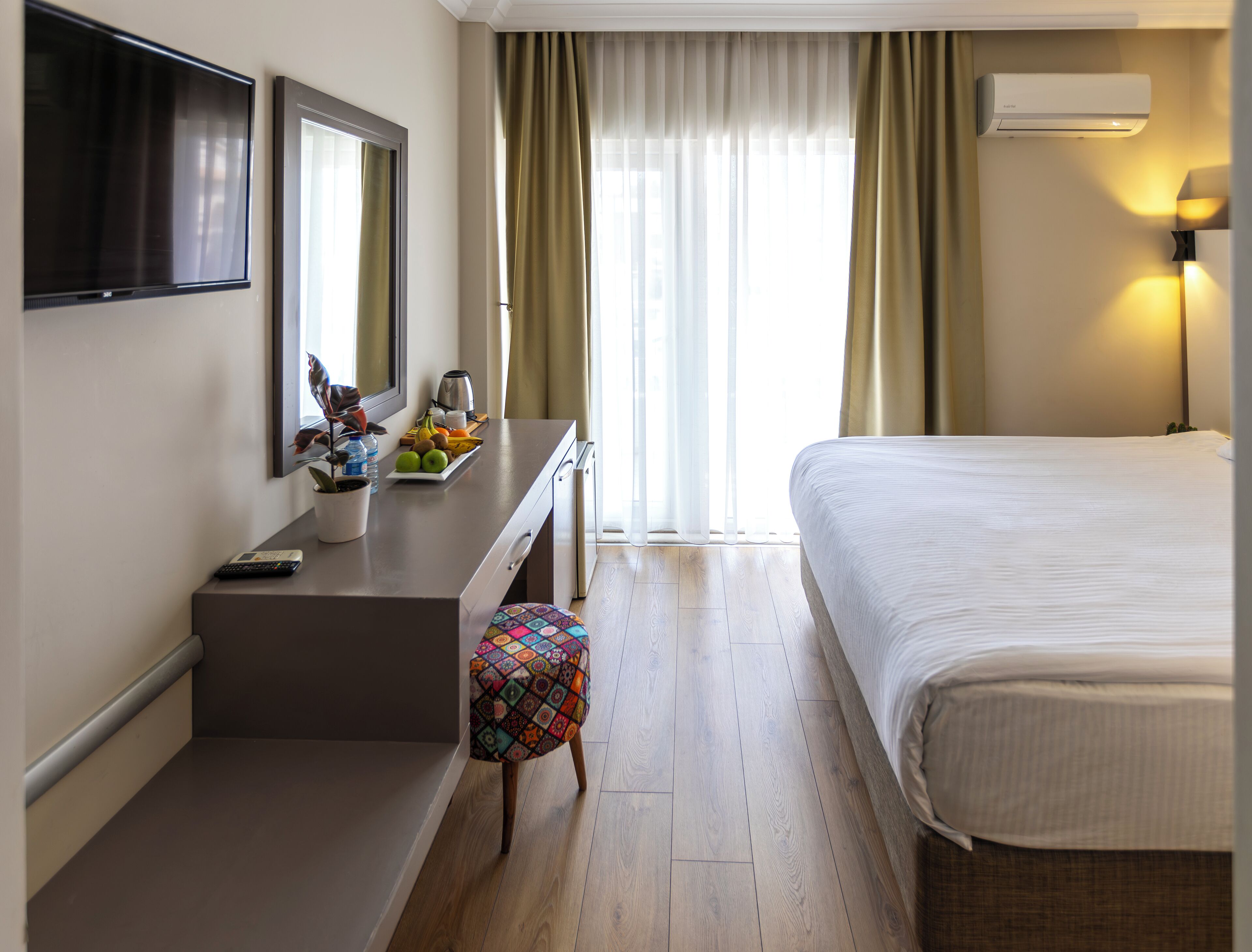 Quarto de hotel com uma cama, uma escrivaninha com cadeira, televisão, janela com cortinas e ar condicionado.