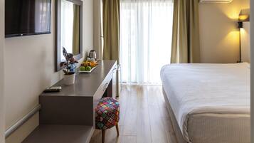 Quarto de hotel com uma cama, uma escrivaninha com cadeira, televisão, janela com cortinas e ar condicionado.